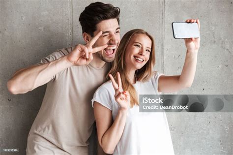 아름다운 부부 남녀가 스마트폰으로 셀카 사진을 찍고 있는 모습 콘크리트 회색 벽 위에 고립된 평화 표지판을 보여주며 실내에서