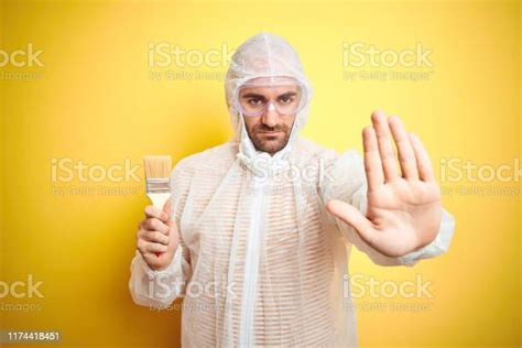 화가 장비를 착용하고 진지하고 자신감있는 표정으로 정지 표지판을하고 열린 손으로 고립 된 노란색 배경 위에 그림 브러시를 들고