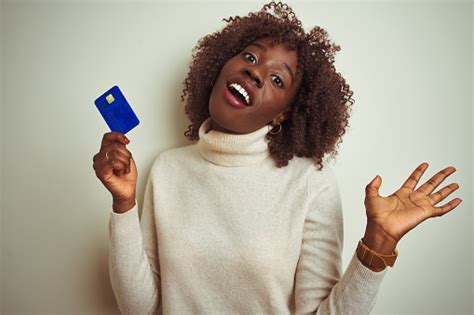 고립된 흰색 배경 위에 서있는 신용 카드를 들고있는 젊은 아프리카 아프로 여성은 매우 행복하고 흥분하며 큰 미소와 손을 들고 비명을 지르는 승리를 축하하는 승자 표정