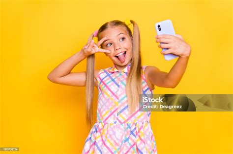 사진 예쁜 작은 아가씨 보유 전화 인플루언서 셀카 블로그 보여주는 V기호 기호 스틱 혀 밖으로 입 착용 체크 무늬 여름 드레스 고립 된 밝은 색상 배경 금발 머리에 대한