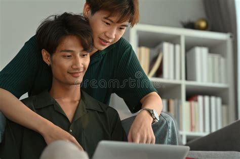 Happy Same Sex Male Couple Embracing And Using Laptop In Living Room