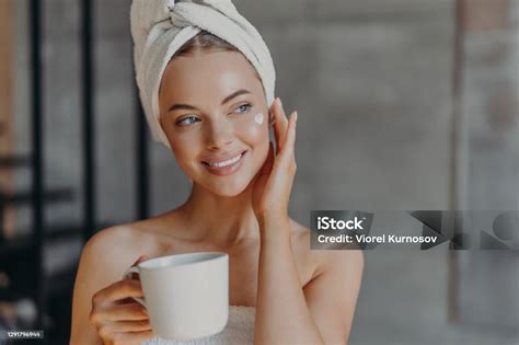 꽤 매력적인 젊은 여성은 부드럽게 얼굴을 터치 얼굴 크림을 적용 최소한의 메이크업을 착용 부드럽게 미소 머리에 포장 된 흰색 목욕 수건을 입고 향기로운 커피를 마시고 집에서