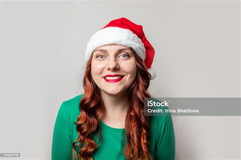 빨간 곱슬 머리를 한 어린 소녀는 빨간 산타 모자를 쓰고 카메라를 보고 회색 배경에 미소를 짓습니다 개념에 대한 스톡 사진 및 기타 이미지 Istock