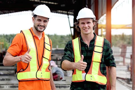 안전 조끼와 헬멧을 착용한 두 명의 행복한 미소 짓는 작업자 엔지니어가 건설 현장에서 검사하는 동안 엄지손가락을 치켜세우고