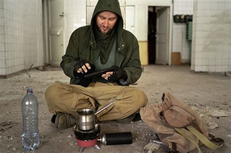 더러운 피곤한 남자가 버려진 방의 바닥에 앉아 가스 버너 위로 손을 따뜻하게하고 저녁 식사를 준비합니다 포스트 묵시록 포스트 묵시록 세계에서 하이킹 35 39세에 대한 스톡