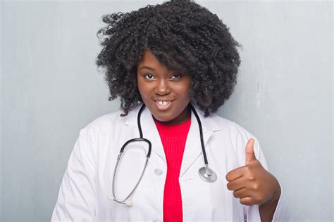 행복 한 엄지손가락 손으로 제스처를 하 고 회색 그런 지 벽에 젊은 아프리카계 미국인 의사 여자 승인 성공을 보여주는 카메라를 보고 하는 식 건강관리와 의술에 대한 스톡