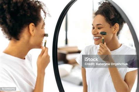 하얀 티셔츠를 입은 아프리카 계 미국인 예쁜 소녀 집에서 거울 앞에 서서 아침 미용 루틴을하고 얼굴 피부를 돌보고 얼굴 옥 롤러를 사용하고 주름을 예방하고 미소 짓습니다