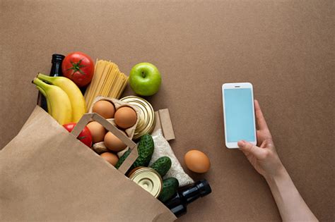 음식용품이 담긴 종이 봉투와 중성 베이지색 배경에 전화로 여성손 음식 배달 검역 기부 코로나 바이러스 개념에 대한 모바일 주문 응용 프로그램 텍스트 모형을 위한 공간 복사