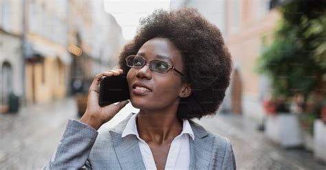 안경과 양복을 입은 매력적인 아프리카 비즈니스 여성이 모바일로 이야기하고 도시 거리에 서 있는 동안 옆을 바라보고 있습니다