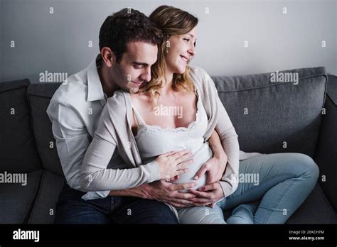 Husband Embracing Pregnant Wife While Sitting On Sofa At Home Stock