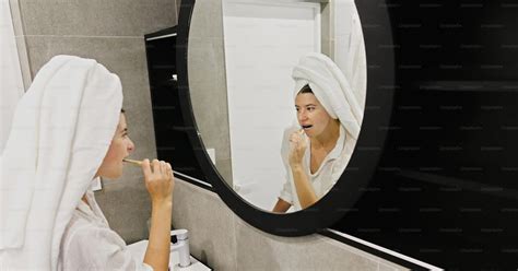 하얀 수건을 쓴 아름다운 젊은 여성이 현대적인 욕실에서 대나무 칫솔과 숯불 치약으로 양치질을 하고 둥근 거울을 보고 있다 치과 위생 폐기물 제로 사진