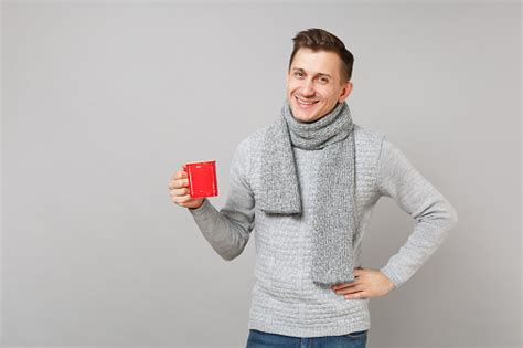 쾌활 한 젊은 남자가 회색 스웨터 스카프 커피 또는 차 스튜디오에 회색 배경에 고립의 빨간 컵을 들고 건강 한 생활 습관 병