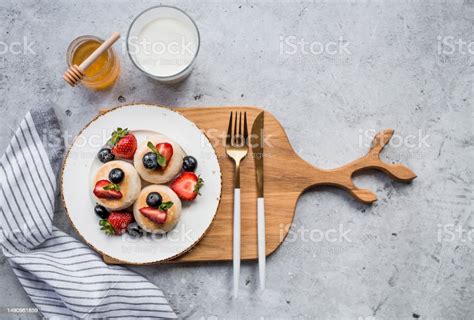 코티지 치즈 팬케이크 신선한 블루베리 딸기 꿀 민트 잎을 곁들인 러시아 치즈 케이크가 회색 콘크리트 테이블 위의 세련된 나무 판에 있습니다 천연 제품 건강하고 맛있는 아침