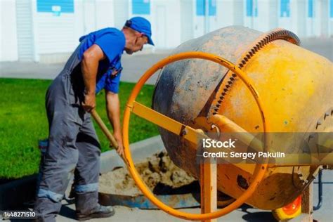 제복을 입은 건설 노동자는 여름날 건설 현장에서 일합니다 노인 벽돌공은 시멘트와 모래를 콘크리트 믹서에 삽질합니다 건물 및 구조물 수리 계약자 석공에 대한 스톡 사진 및