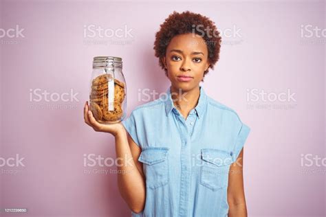 분홍색 고립 된 배경 위에 초콜릿 칩 쿠키 항아리를 들고있는 아프리카 계 미국인 여성은 스마트 한 얼굴에 자신감있는 표정을