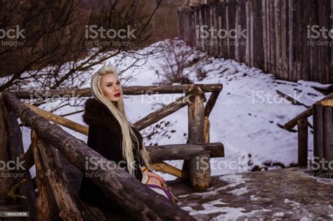 모피 칼라가 달린 전통 옷을 입은 아름다운 스칸디나비아 전사 생강 여성의 야외 초상화와 손에 칼 나무 바이킹 빌리지가 배경에 보이는 모습 20 24세에 대한 스톡 사진 및