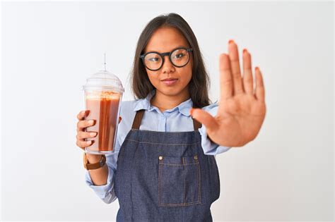 안경을 쓰고 있는 중국 여성은 고립된 흰색 배경 위에 스무디를 마시고 진지하고 자신감 있는 표정으로 정지 표지판을 하고 방어 제스처를 취합니다 건강한 생활방식에 대한 스톡