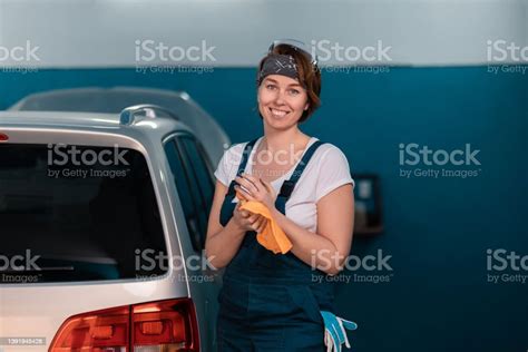 푸른 작업복을 입은 행복한 백인 젊은 여성이 헝겊으로 손을 흔들고 있는 모습 배경에있는 자동차와 워크샵 자동차 수리점에서 일하기 기계 부분에 대한 스톡 사진 및 기타 이미지