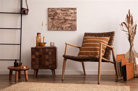 모의 포스터 프레임 나무 찬장 말린 꽃이 있는 유리 꽃병 베이지색 카펫 갈색 안락의자 및 개인 액세서리가 있는 거실 공간의 따뜻하고 아늑한 인테리어 가정 장식 템플렛 0명에