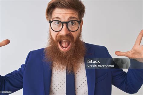 고립된 흰색 배경 위에 양복과 안경을 착용한 젊은 빨간 머리 아일랜드 사업가가 미친 듯이 축하하고 팔을 들고 눈을 뜨고 흥분을 일으키며 성공을 축하했습니다 우승자 컨셉