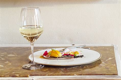 지중해 생 농어 구이 생선 필레 찐 감자 검은 올리브 태양 말린 분말 무 캐러멜레드 양파와 레몬 처트니 에 포르셀리안 고급 이탈리아 디자인 화이트 접시 루가나 화이트 와인