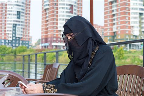 검은 옷을 입은 이슬람 여성이 손에 전화기를 들고 여름 카페에 앉아 있습니다 Brand Name Smart Phone에 대한 스톡 사진 및 기타 이미지 Brand