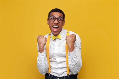멜빵과 나비 넥타이를 매고 흰 셔츠를 입은 젊은 아프리카계 미국인 남자가 노란색 배경에 승리를 기뻐한다 긍정적인 감정 표현에