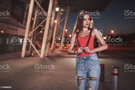 그녀의 어깨 뒤에 배낭과 젊은 아름 다운 소녀는 따뜻한 여름 저녁에 공항 또는 기차역 근처 거리에 서 있다 그녀는 그냥 도착 하 고 택시 또는 그녀의 친구를 기다리고 가방에