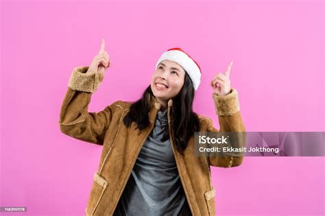 초상화 여성들은 두꺼운 천을 입고 크리스마스 모자를 쓰고 몸을 따뜻하게 유지하고 머리를 위로 올리고 똑바로 서서 검지 손가락을 위로 향하게 하고 분홍색 배경에 고립된 실내