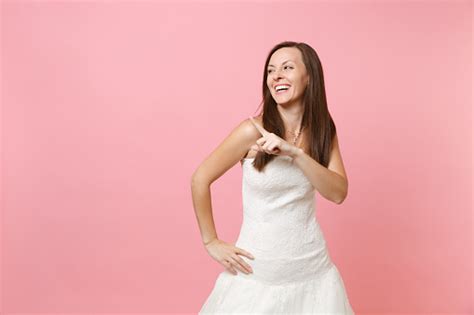 하얀 웨딩 드레스 서 포인팅 검지 손가락 보고 옆으로 파스텔 핑크 배경에 고립에서 즐거운 신부 여자 웃음의 초상화 결혼식 축 하 개념입니다 광고에 대 한 공간을 복사 합니다