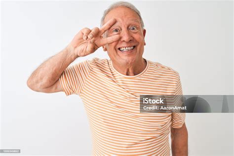고립 된 흰색 배경 위에 서있는 줄무늬 티셔츠를 입은 선임 회색 머리의 남자가 얼굴에 손가락으로 평화 기호를하고 승리를 보여주는 쾌활한 미소를 지었습니다 T 셔츠에 대한