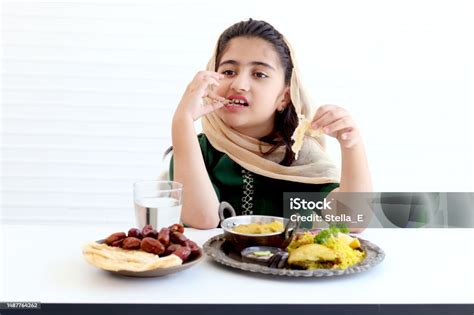 부엌 식탁에 앉아 아름다운 눈을 가진 사랑스러운 웃는 파키스탄 무슬림 소녀 히잡과 전통 의상을 입은 아이는 하얀 배경에 손으로 전통적인 이슬람 할랄 음식을 먹는 것을 즐긴다