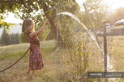Girl In Dress Watering Tree With Hose In Sunny Summer Yard Looking Down Garden Stock Photo