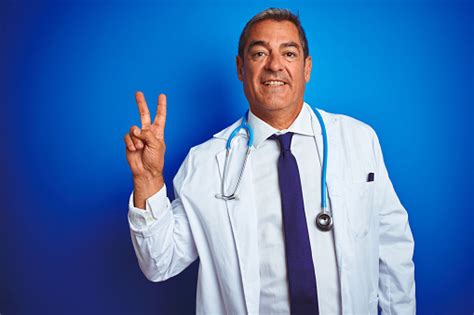 고립 된 파란색 배경 위에 청진기를 입은 잘 생긴 중년 의사 남자가 승리의 표지판을하고있는 카메라에서 행복한 얼굴 윙크와 미소 2번 건강관리와 의술에 대한 스톡 사진 및