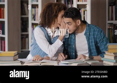 Two Students Of Classmate Sitting At Desk In Lesson Doing Homework In Library Girl Whispers In