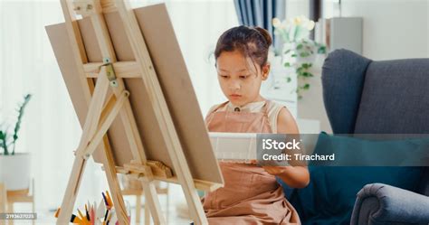 앞치마를 두른 아시아 유아 소녀는 집에서 주말에 추상화를 그리는 데 초점을 맞춘 페인트 브러시를 사용하여 그림 캔버스 앞의 소파 의자에 앉아 있습니다 아이를 위한 창의적인