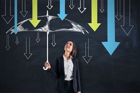 매력적인 젊은 유럽 사업가는 칠판 벽 배경에 화려한 하향 화살표로부터 자신을 보호하기 위해 그려진 우산 스케치를 들고 금융 투자 및 보험 개념 개념에 대한 스톡 사진 및