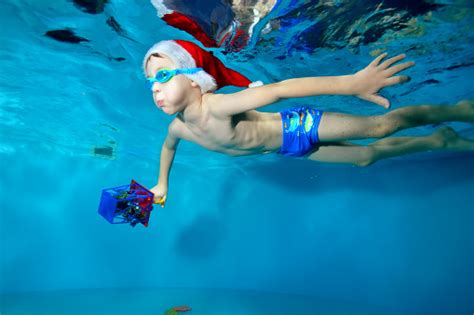 빨간 산타 모자를 쓴 어린 소년이 손에 크리스마스 선물을 들고 어린이 수영장에서 수중 수영을 하고 팔을 뻗습니다 크리스마스 축하 초상화 개념 12월에 대한 스톡 사진 및
