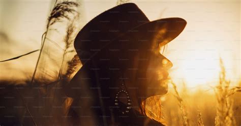 가을 들판의 따뜻한 일몰 빛 속에서 풀밭에 서 있는 모자를 쓴 아름다운 세련된 여성의 실루엣 고요한 분위기의 순간 시골에서 저녁을 즐기는 세련된 젊은 여성 사진