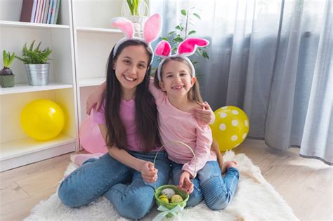 분홍색 티셔츠와 토끼 귀를 입은 두 명의 웃는 자매 소녀가 풍선으로 장식된 방에서 부활절 휴가를 축하하는 바구니에 달걀을 수집합니다 2명에 대한 스톡 사진 및 기타 이미지