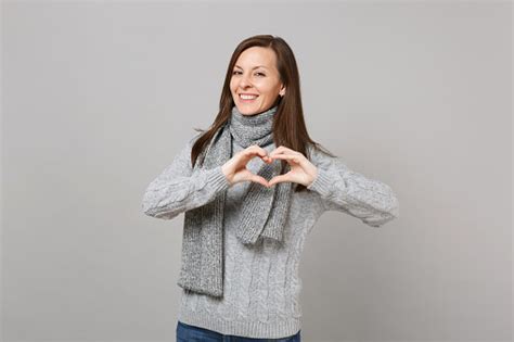 회색 스웨터에 젊은 여자를 웃 고 회색 벽 배경에 고립 손으로 표시 모양 심장 스카프 건강 패션 라이프 스타일 사람들이 진심으 감정 추운 계절 개념 복사 공간을 비웃는 다