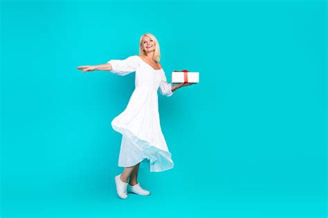 흐르는 듯한 흰색 드레스를 입은 우아한 금발 여성이 밝은 청록색 배경의 선물 상자를 들고 행복과 매력을 표현합니다 45 49세에 대한 스톡 사진 및 기타 이미지 Istock