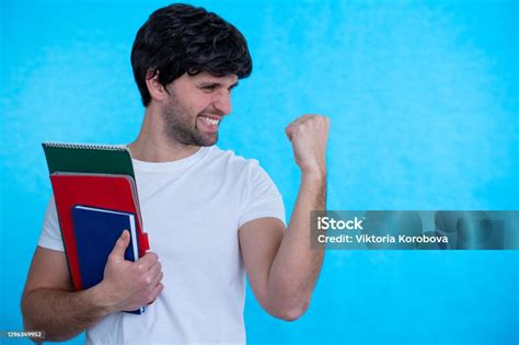승리를 축하하는 좋은 기분을 가진 주먹을 들고 손을 들고 있는 남자 시험을 통과하고 작업을 마치고 파란 배경 위에 서 있는 세 권의 다채로운 카피 북을 들고 있는 남자