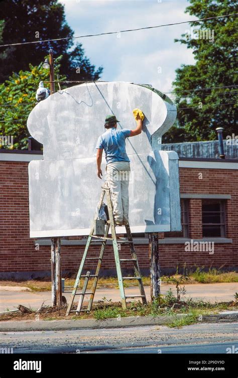 Painter On Step Ladder Preps The Blank Surface Of An Outdoor Sign Shaped Like A Slice Of Bread
