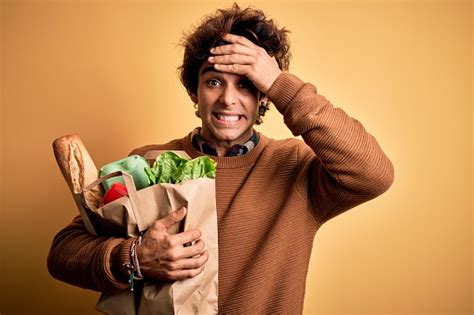 고립된 노란색 배경 위에 음식이 서있는 종이 가방을 들고있는 젊은 잘생긴 남자는 머리에 손으로 스트레스를 받고 수치심과 깜짝 얼굴에 충격을 받았으며 화가 나고 좌절했습니다