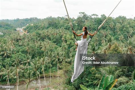 기차를 타고 긴 흰색 드레스를 입은 아름다운 여성 그네를 타고 배경에는 열대 우림과 야자수가 있습니다 공간을 복사합니다 후면 보기 경관에 대한 스톡 사진 및 기타 이미지