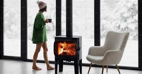 겨울철 자연에 있는 현대 주택의 창문 근처 벽난로 옆에 컵을 들고 서 있는 여자 집에서 겨울 분위기와 편안함의 개념 산속의 오두막에서 레크리에이션에 대한 아이디어 사진