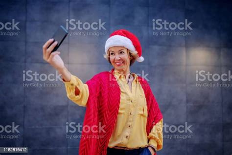 매력적인 미소 백인 선임 금발 의 여자 와 산타 모자 머리에 포즈와 셀카를 복용 하는 동안 벽 앞에 서 야외 12월에 대한 스톡 사진 및 기타 이미지 Istock