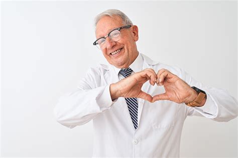 고립 된 흰색 배경 위에 서있는 코트를 입은 선임 회색 머리 과학자 남자는 손으로 심장 기호와 모양을 보여주는 사랑에 미소