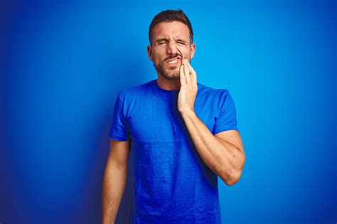 치통이나 치아 질환으로 인해 고통스러운 표정으로 입을 만지는 파란색 고립 된 배경 위에 캐주얼 한 티셔츠를 입은 젊은 잘생긴 남자 치과 의사 개념 T 셔츠에 대한 스톡 사진